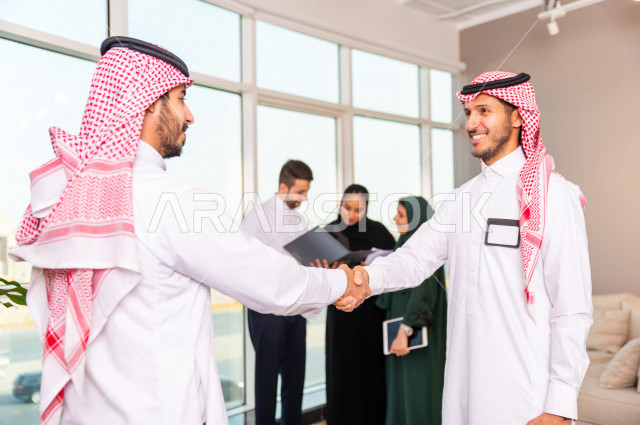 رجل اعمال سعودي خليجي يصافح شريكه في العمل ، و بالخلفية مجموعة من