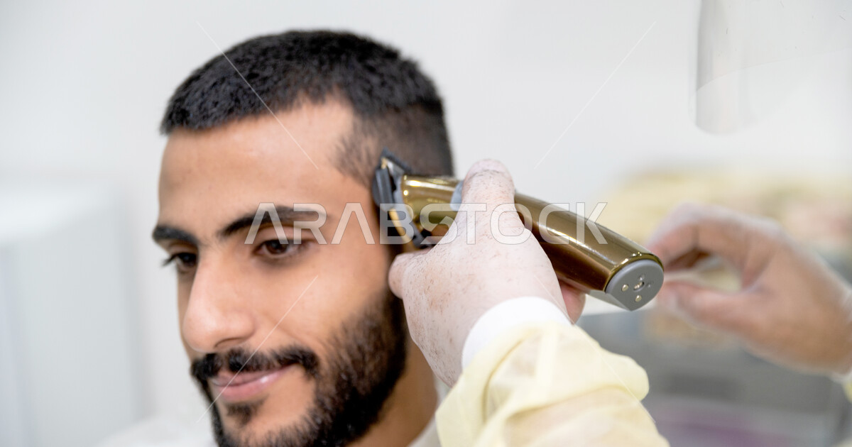 A Saudi Arabian Gulf man visits a barber shop. The barber uses the ...