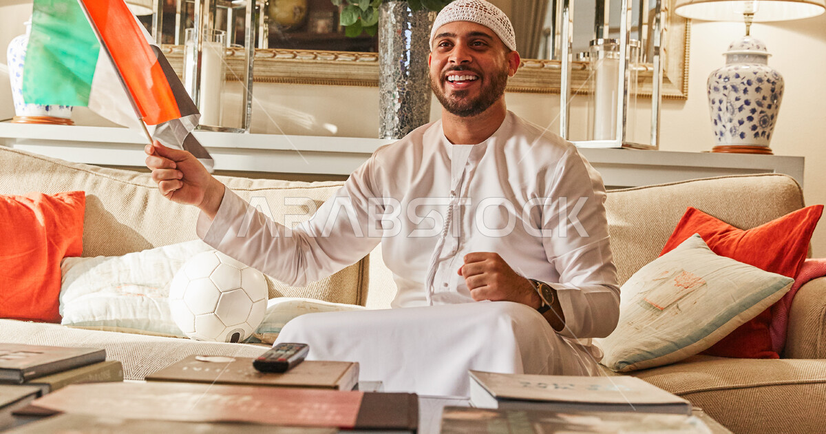 Watching a football match, a smiling Emirati Gulf Arab man wearing a ...