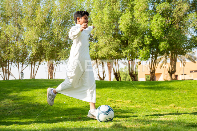 Playing football, a Saudi Gulf Arab boy wearing traditional clothing ...