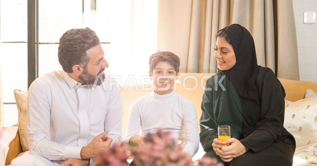 أسرة خليجية صغيرة وسعيدة تجلس على الأريكة في المنزل ، الاستمتاع بقضاء