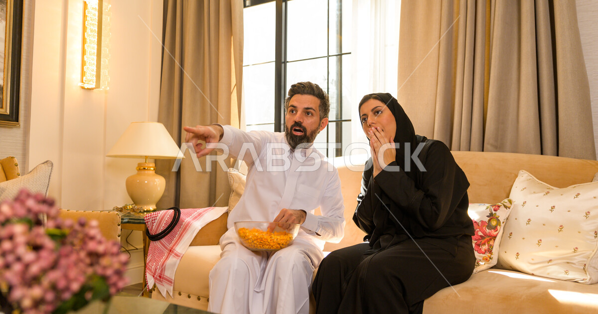 زوجان خليجيان سعيدان يجلسان على الأريكة في المنزل ، الاستمتاع بقضاء