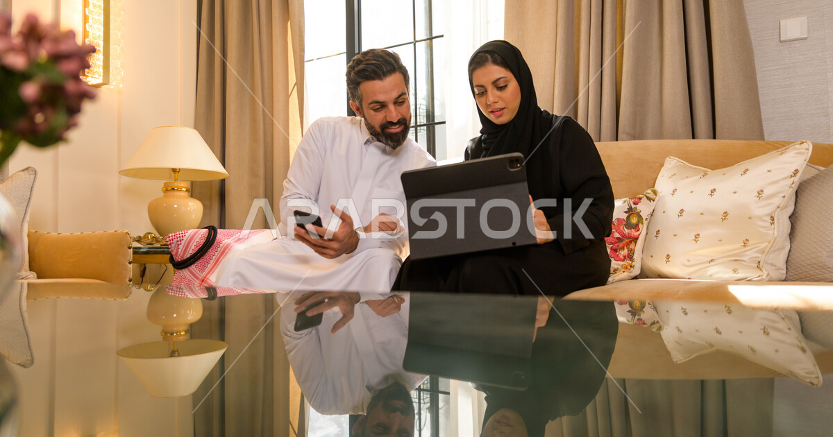 زوجان خليجيان سعيدان يجلسان على الأريكة في المنزل ، يستخدمان الابتوب