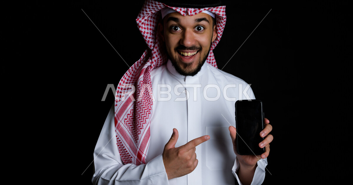 Portrait of a young saudi Gulf Arab pointing to cell phone in amazement ...