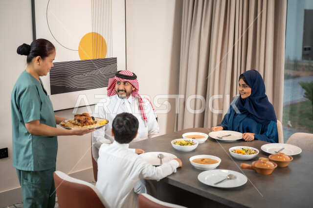 A Saudi Gulf Arab family enjoys eating, a foreign worker preparing the ...