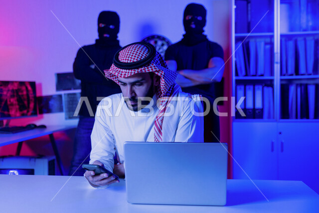 A Saudi Arabian Gulf man sitting in front of a laptop, using a mobile ...