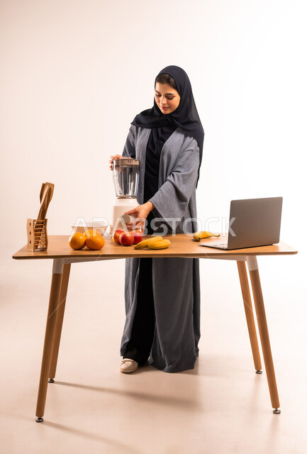 Using laptop, portrait of veiled Saudi Arabian Gulf woman making juice ...