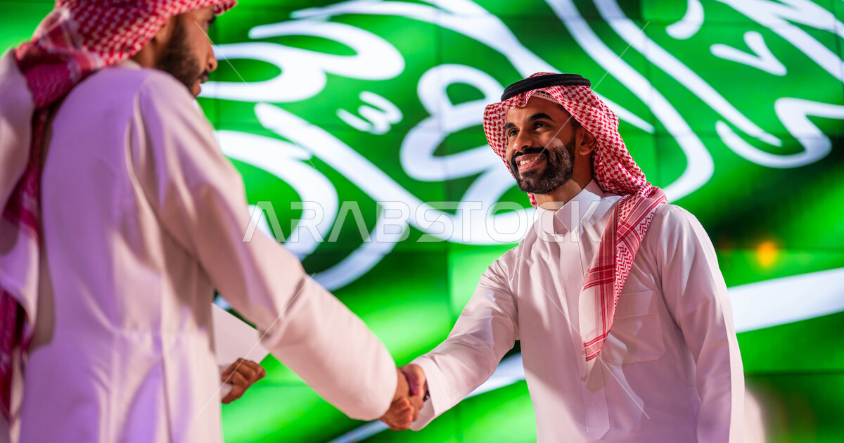 Two Saudi Arabian Gulf men shake hands in front of the background of ...