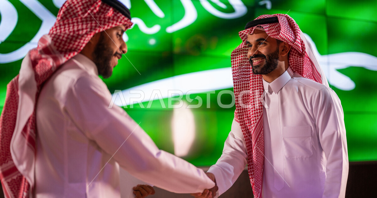 Two Saudi Arabian Gulf men shake hands in front of the background of ...