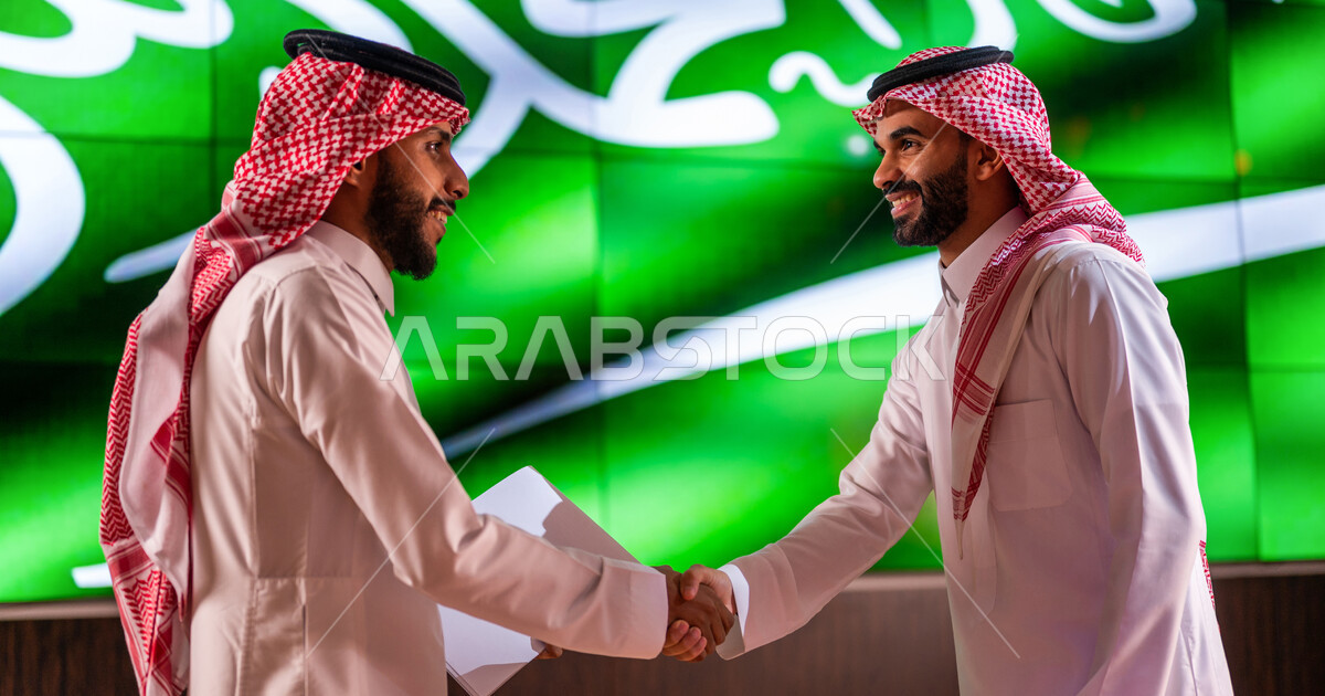 Two Saudi Arabian Gulf men shake hands in front of the background of ...