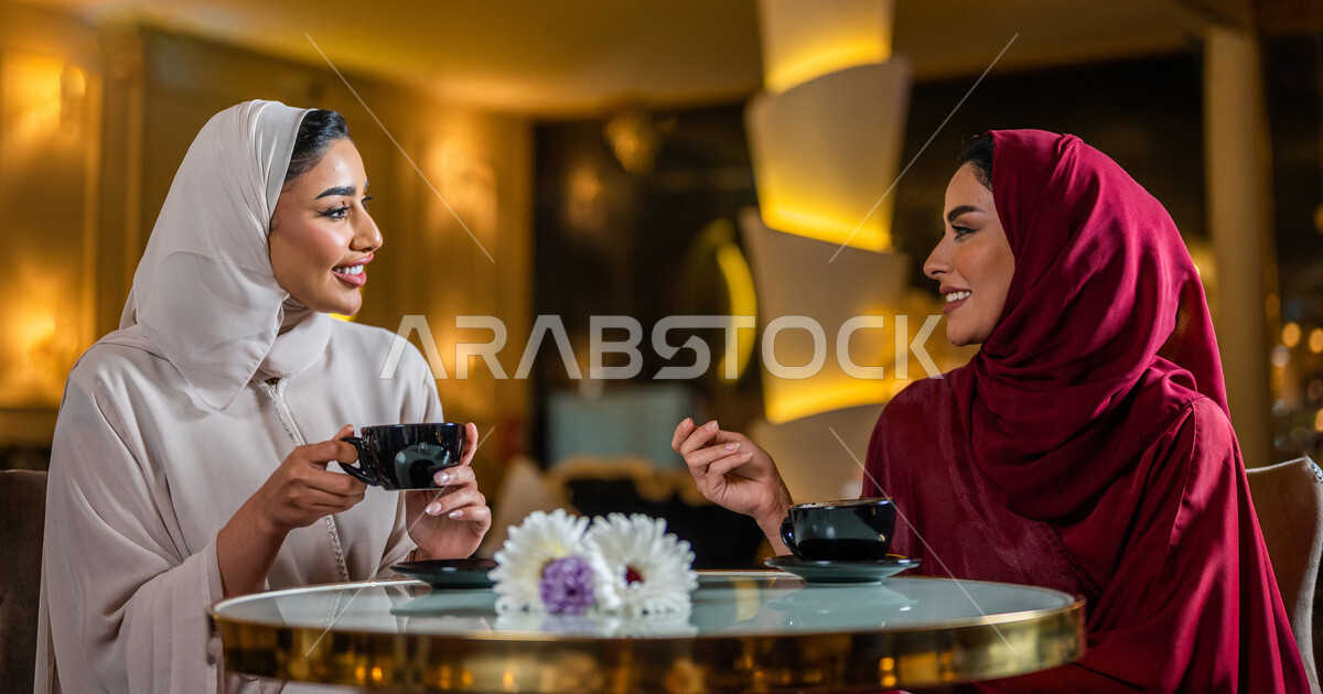 Two smiling Saudi Arabian Gulf women sitting in a café drinking a cup ...