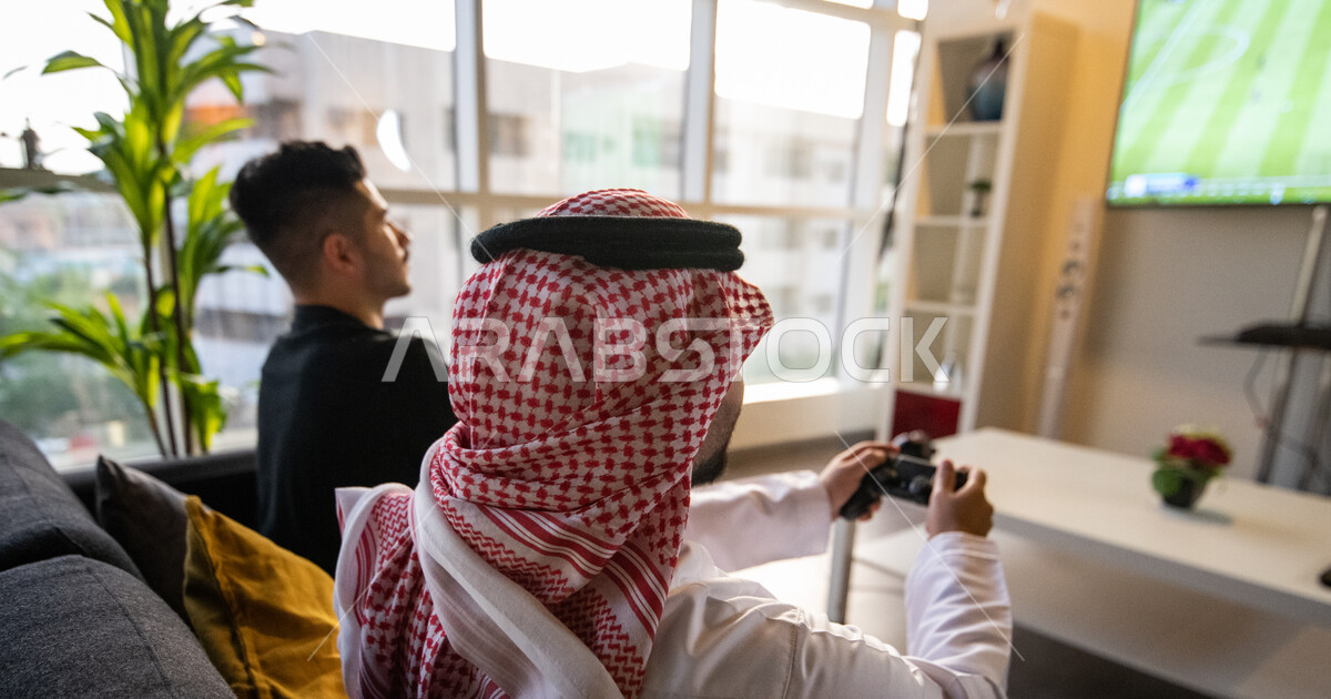Two Saudi Arabian Gulf youths holding a joystick and playing video ...