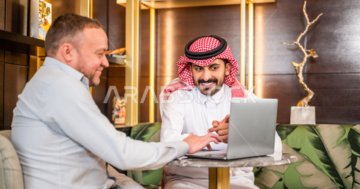Two Saudi Arabian Gulf businessmen using laptop computers and discussing work, business partners ...