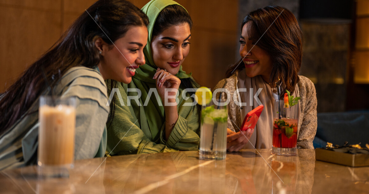 Saudi Arabian Gulf Arab female friends sitting in a café, using a ...