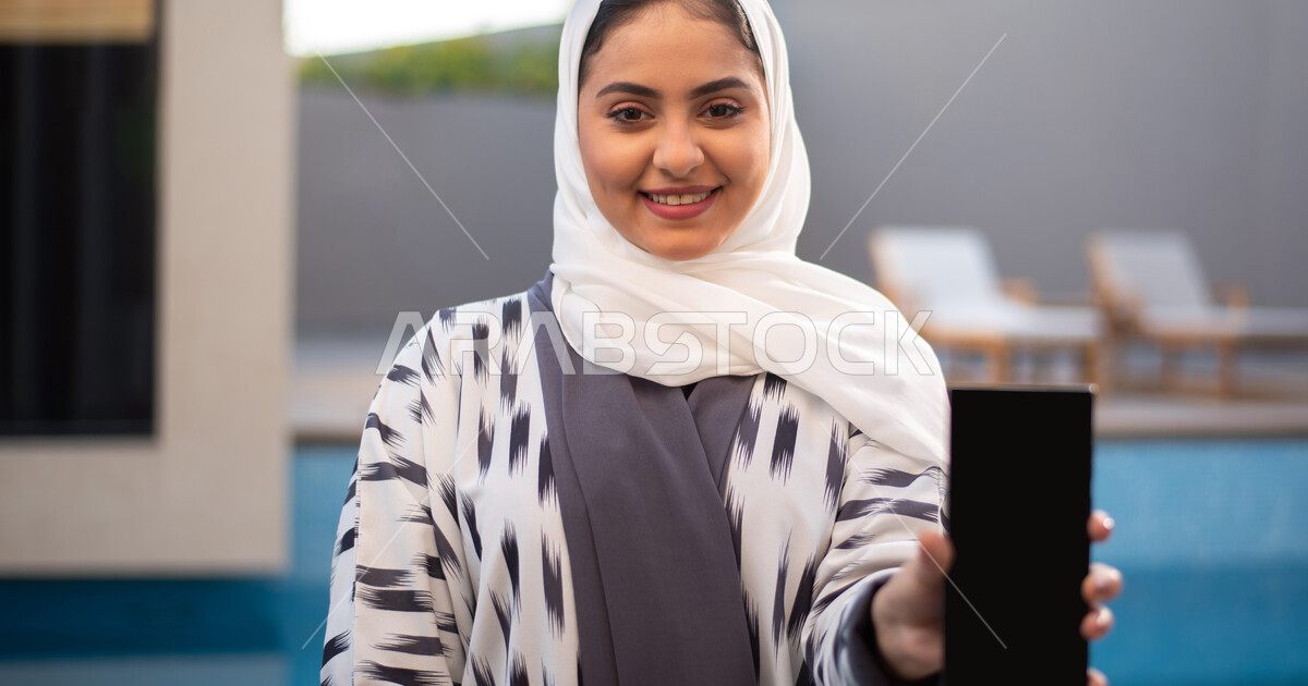 صورة مقربة لمرأة عربية خليجية سعودية ، مبتسمة تنظر إلى الكاميرا ، عرض شاشة سوداء بإستخدام الهاتف ...