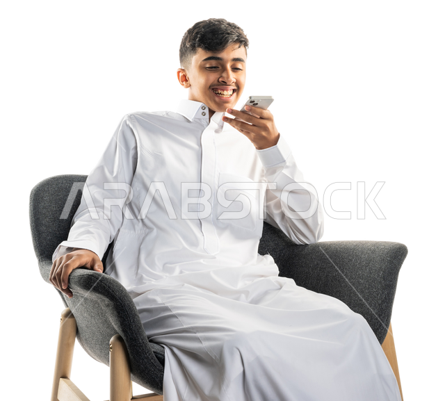 Portrait of a smiling Saudi Arabian Gulf boy, sitting on a chair, using ...