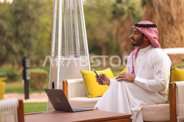 A Saudi Arabian Gulf man browsing a mobile phone, using a laptop ...