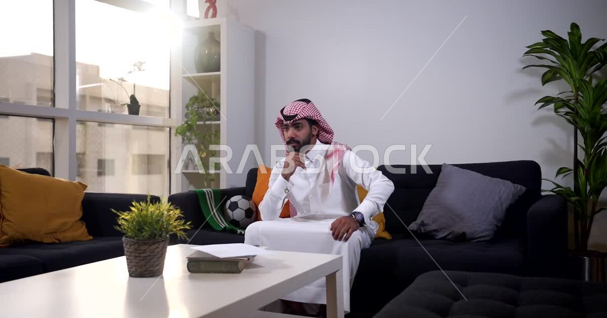 A young Saudi Arab fan for his team, sitting on a sofa in the living ...