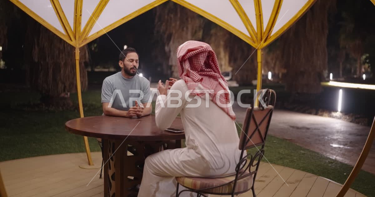 Two Saudi Arabian Gulf men sitting in a public park in the open air at ...